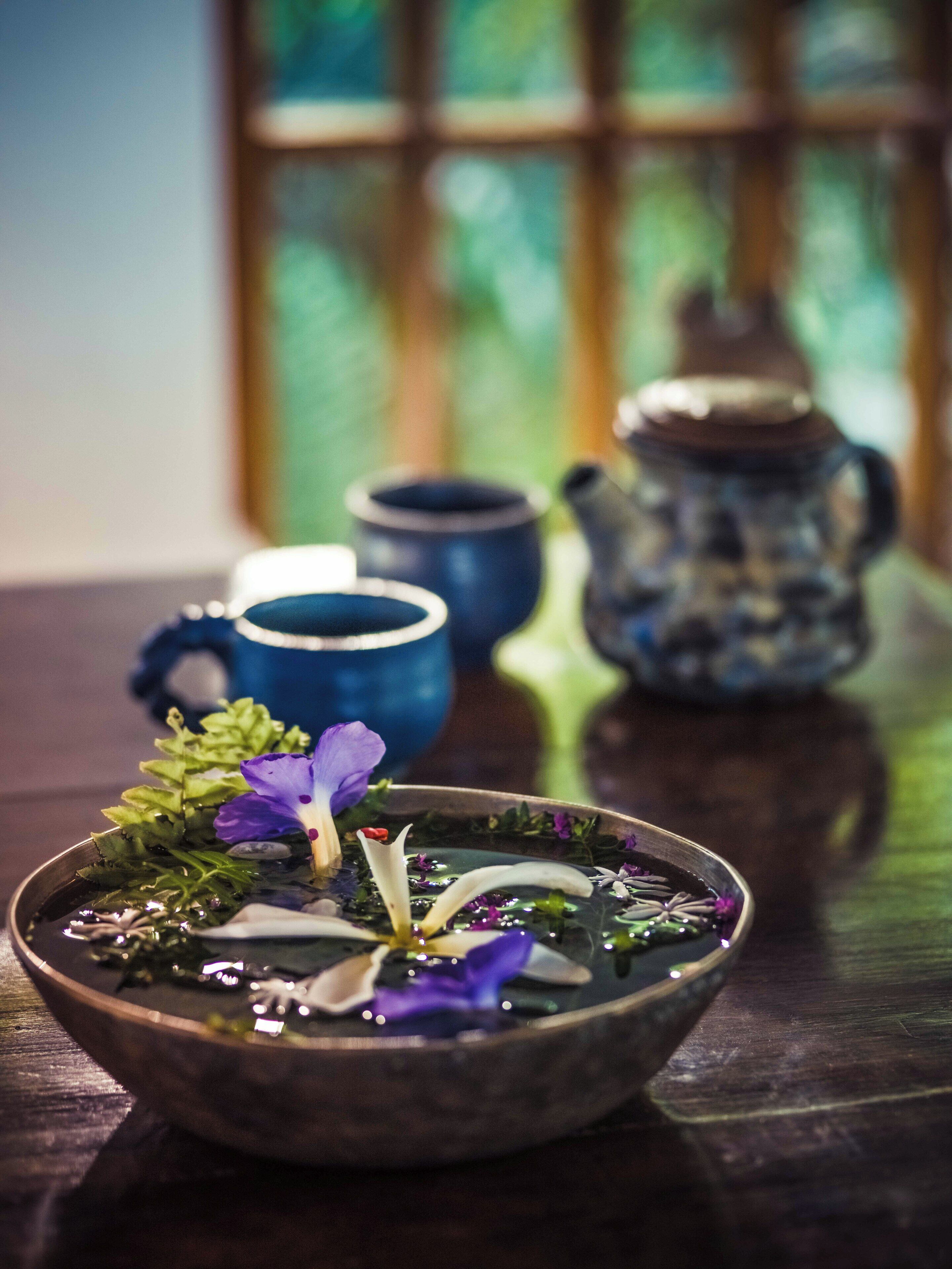 Uma tigela de sopa com ervas e flores, uma chávena azul e um bule sobre uma mesa de madeira.
