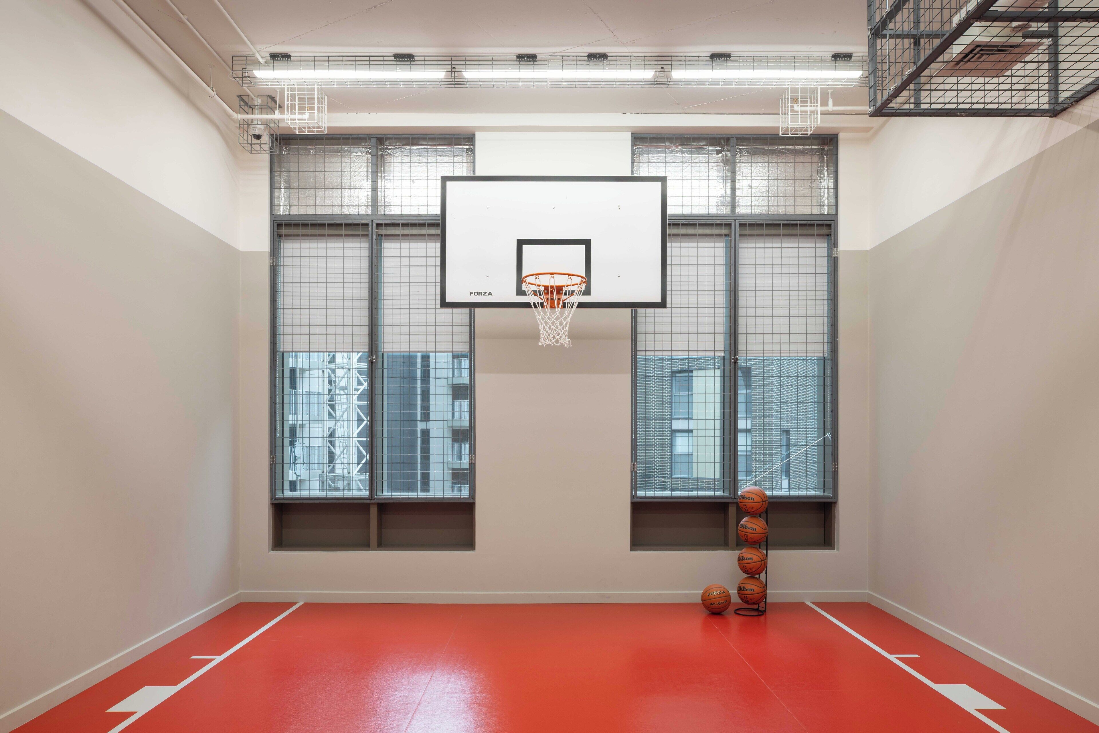Uma quadra de basquete interna com uma cesta, janelas e uma fileira de bolas de basquete.