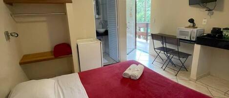 Quarto com cama, manta vermelha, pequena cozinha e televisão, além de porta que dá para uma varanda.