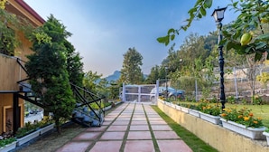 Uma trilha pavimentada conduz a um portão fechado, cercada por vegetação e um edifício com telhado inclinado.