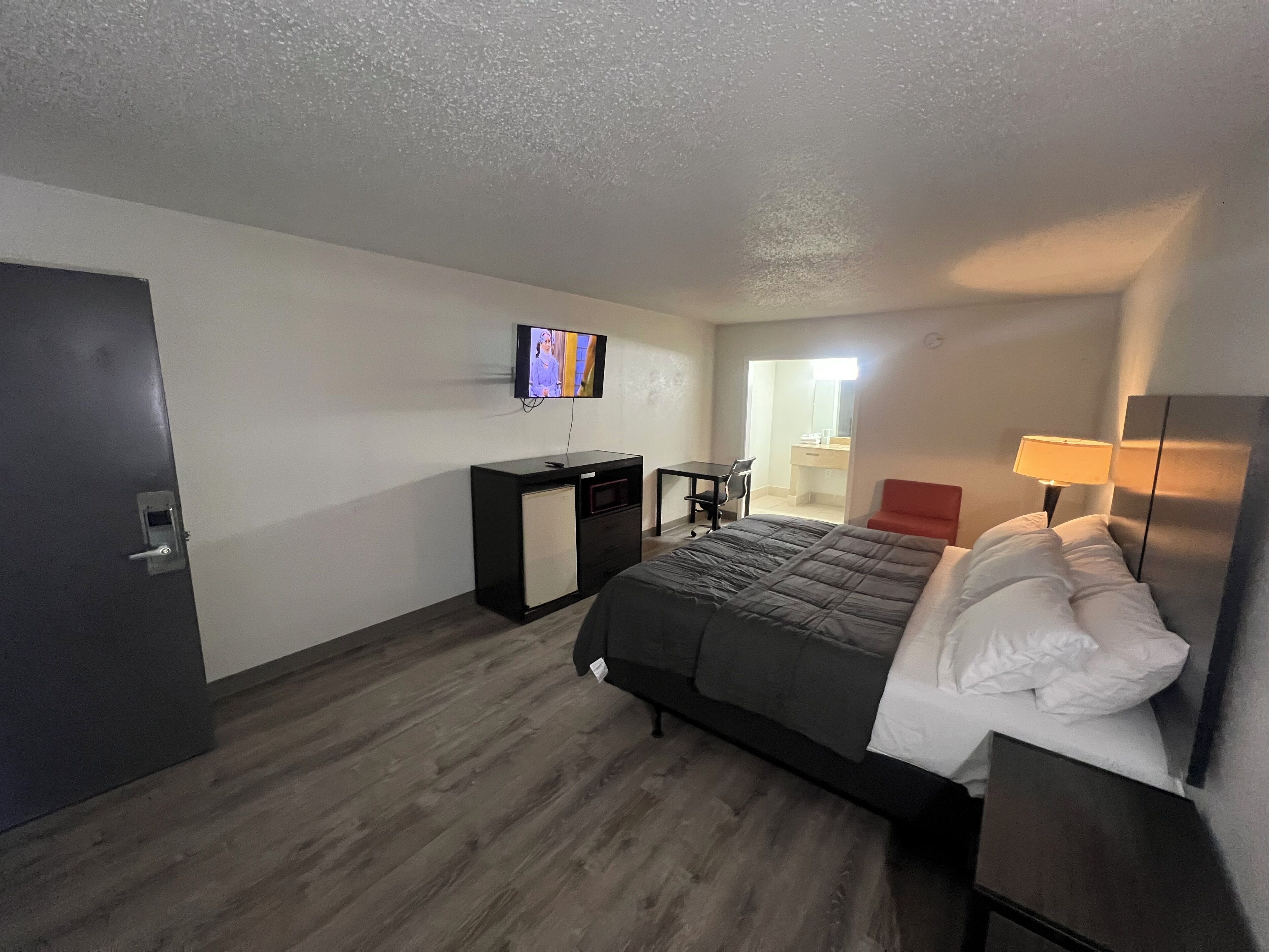 Quarto de hotel com cama, escrivaninha, cadeira, televisão e banheiro visível por uma porta aberta.