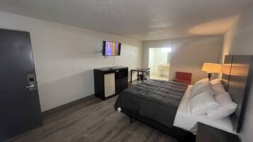 Quarto de hotel com cama, escrivaninha, cadeira, televisão e banheiro visível por uma porta aberta.