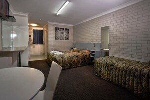 Quarto de hotel pequeno com cama, escrivaninha, cadeira, espelho e uma obra de arte na parede.