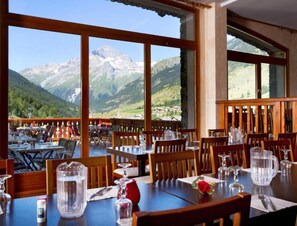 Restaurante com mesas preparadas para refeições, grandes janelas com vista para montanhas e um deck de madeira.