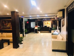 Uma área de recepção de hotel com balcão, espaço para sentar e plantas em vasos.
