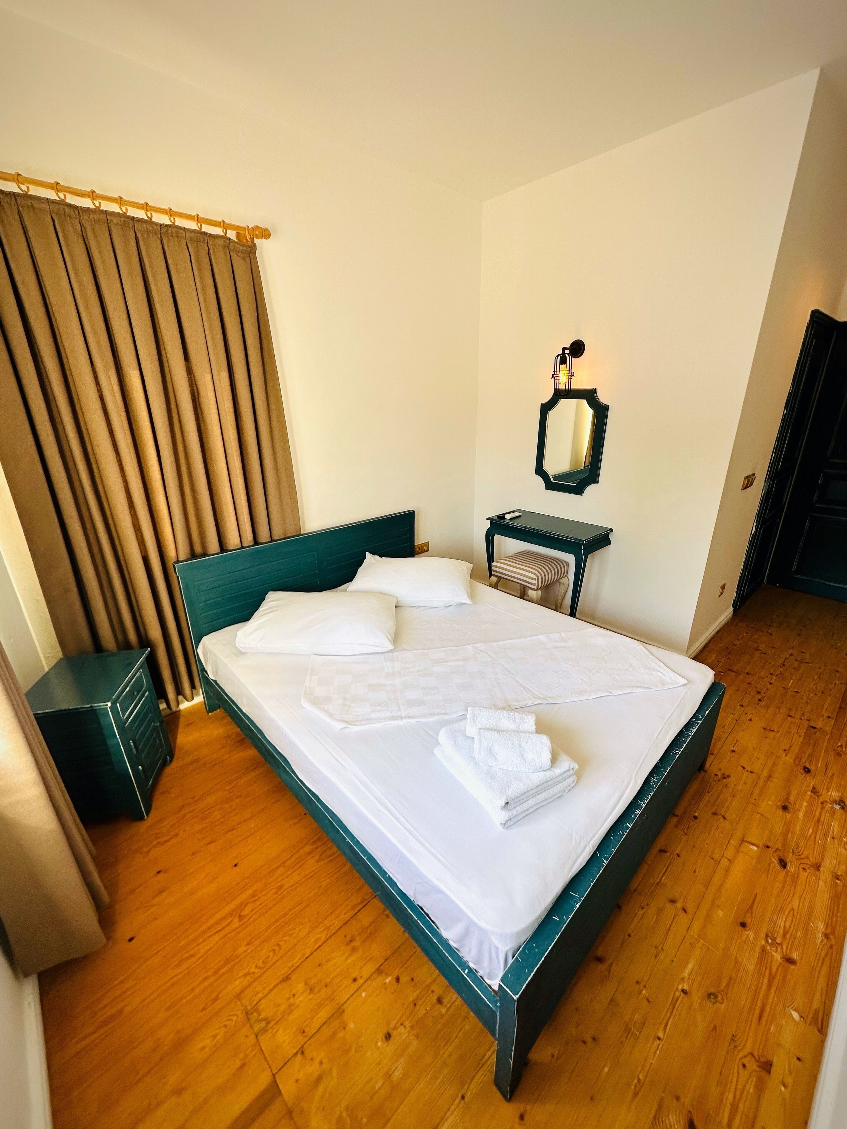 Uma cama bem arrumada com lençóis brancos, um criado-mudo de madeira com abajur e piso de madeira.