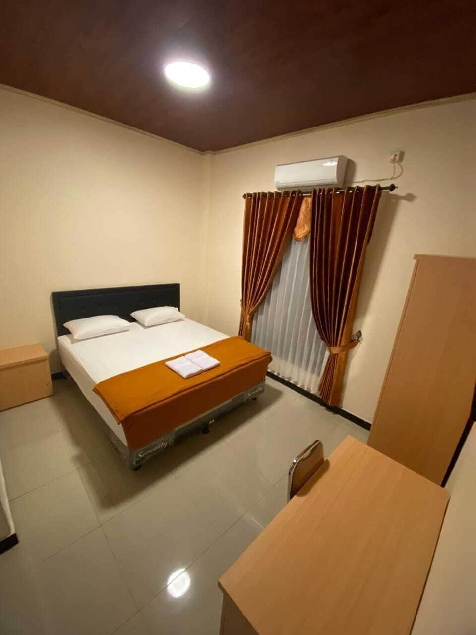 Quarto de hotel com cama, ar condicionado, cortinas, secretária de madeira e guarda-roupa.