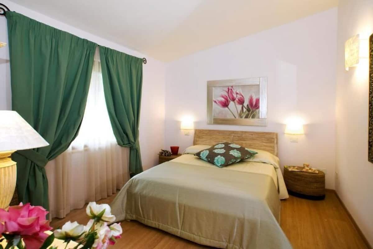 Quarto com cama, cortinas verdes, mesa de cabeceira, abajur, quadro emoldurado e espelho.
