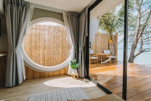 Quarto com uma grande janela circular, parede de bambu, uma pequena mesa com uma planta e vista para o mar.