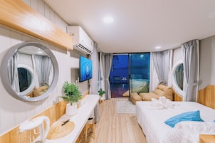 Quarto de hotel moderno com piso de madeira, espelho redondo, televisão, cama com almofadas, mesa pequena com planta e vista para o exterior.