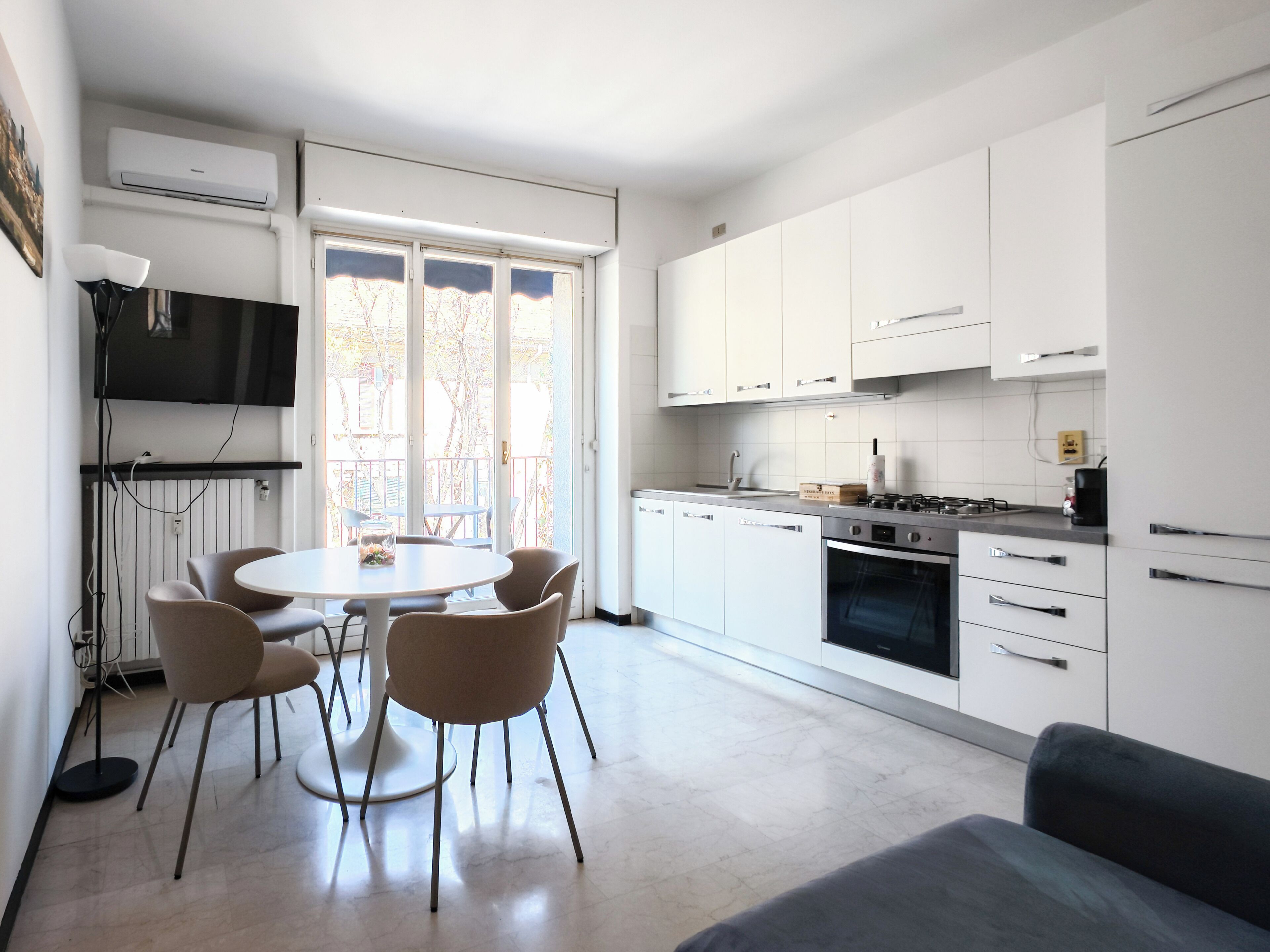 Cozinha moderna com armários brancos, área de jantar com mesa e cadeiras redondas, e uma televisão de tela plana na parede.