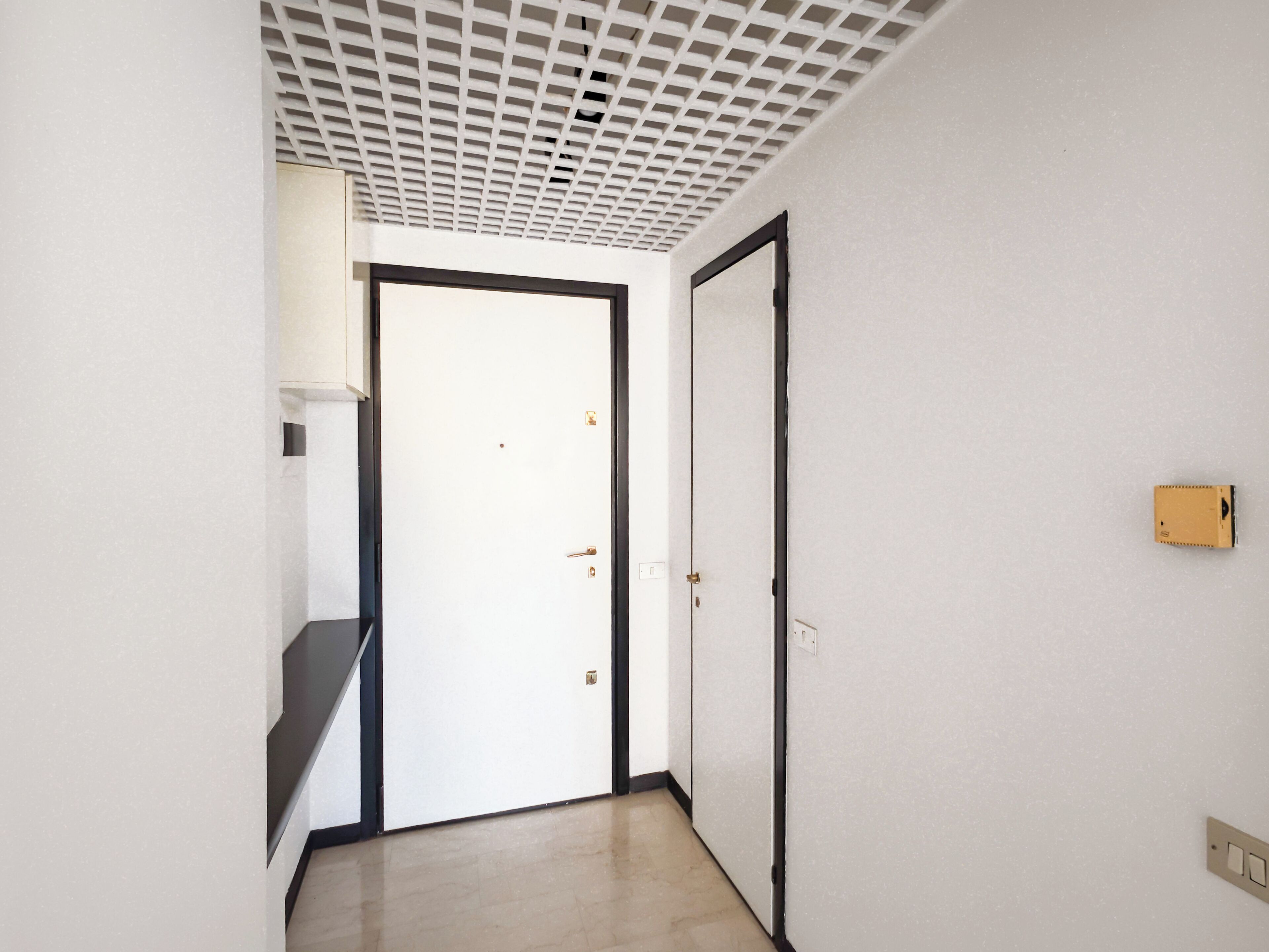 Um corredor com uma porta branca, uma porta com moldura preta e um interruptor na parede.