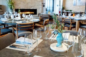 Um restaurante com mesas preparadas para refeições, uma lareira ao fundo e um vaso azul com folhagem sobre a mesa.