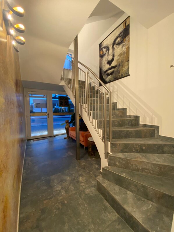 Uma escadaria com corrimão de metal, uma grande pintura abstrata na parede e vista para um quarto com um sofá e uma estátua.