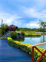 Uma trilha leva a um lago com uma grande escultura decorativa, cercada por vegetação e um céu claro.
