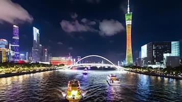 Paisagem urbana noturna com edifícios iluminados, uma ponte e barcos em um rio.