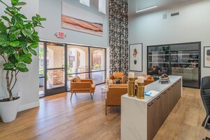 Uma área de recepção moderna com balcão de madeira, assentos confortáveis, uma grande planta em vaso e vista para o pátio externo através de portas de vidro.