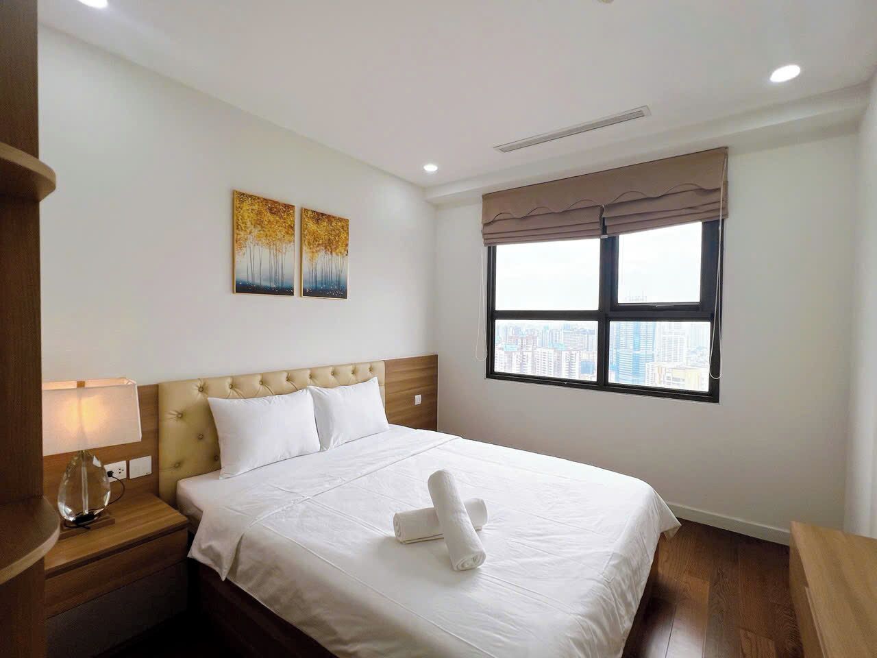 Quarto de hotel com cama, mesa de cabeceira, luminária, janela com vista para prédios e obras de arte na parede.