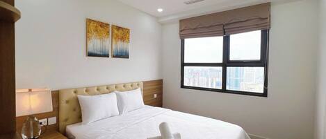 Quarto de hotel com cama, mesa de cabeceira, luminária, janela com vista para prédios e obras de arte na parede.