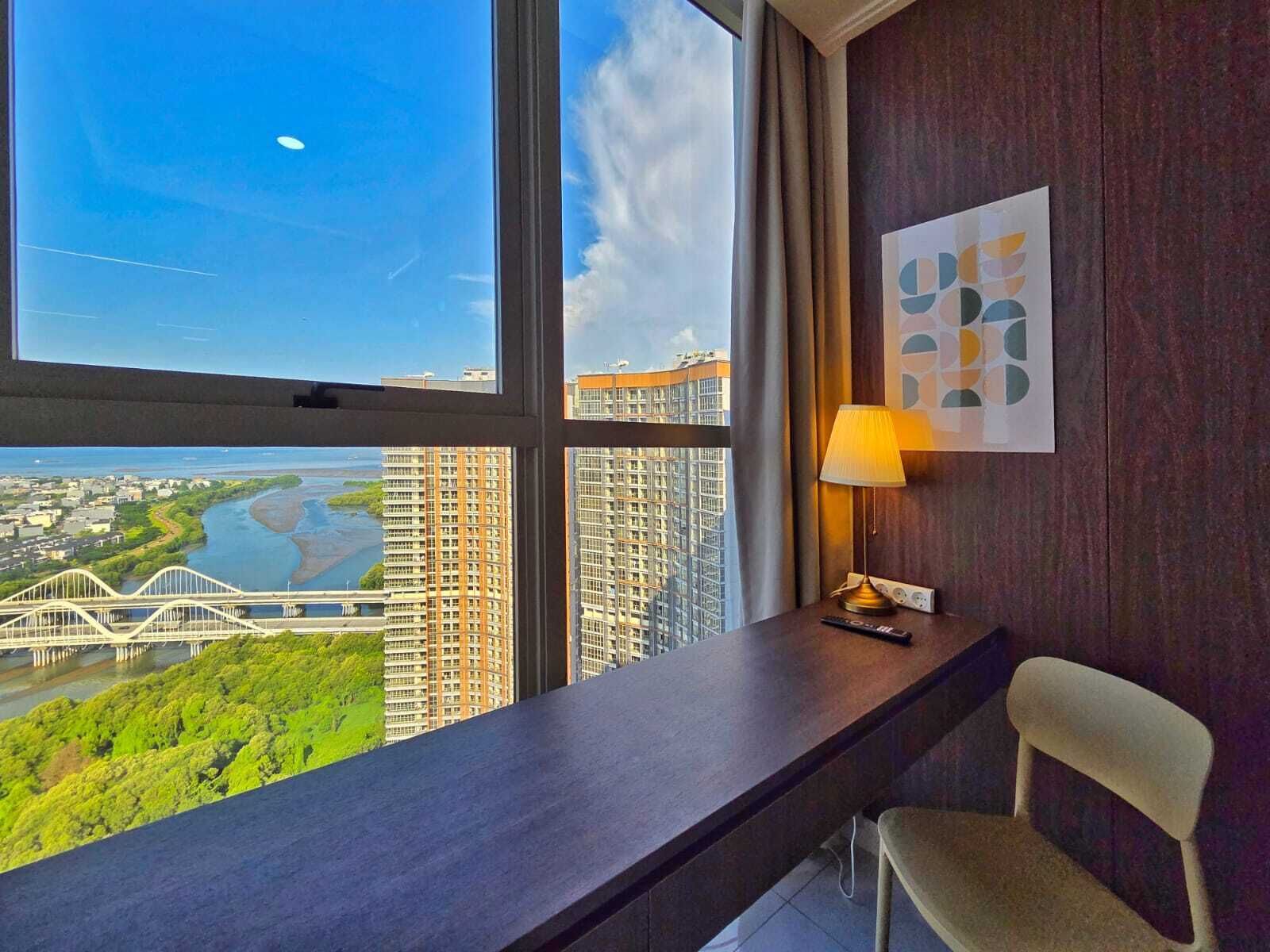 Quarto de hotel com escrivaninha, cadeira, luminária e uma grande janela com vista para uma ponte e áreas verdes.