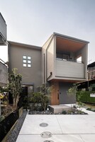Um edifício moderno de dois andares com varanda, um pequeno jardim com plantas e um caminho de concreto que leva à entrada.