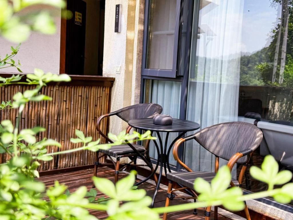 Um pátio com mesa redonda e duas cadeiras, cerca de bambu e janela com vista para vegetação.