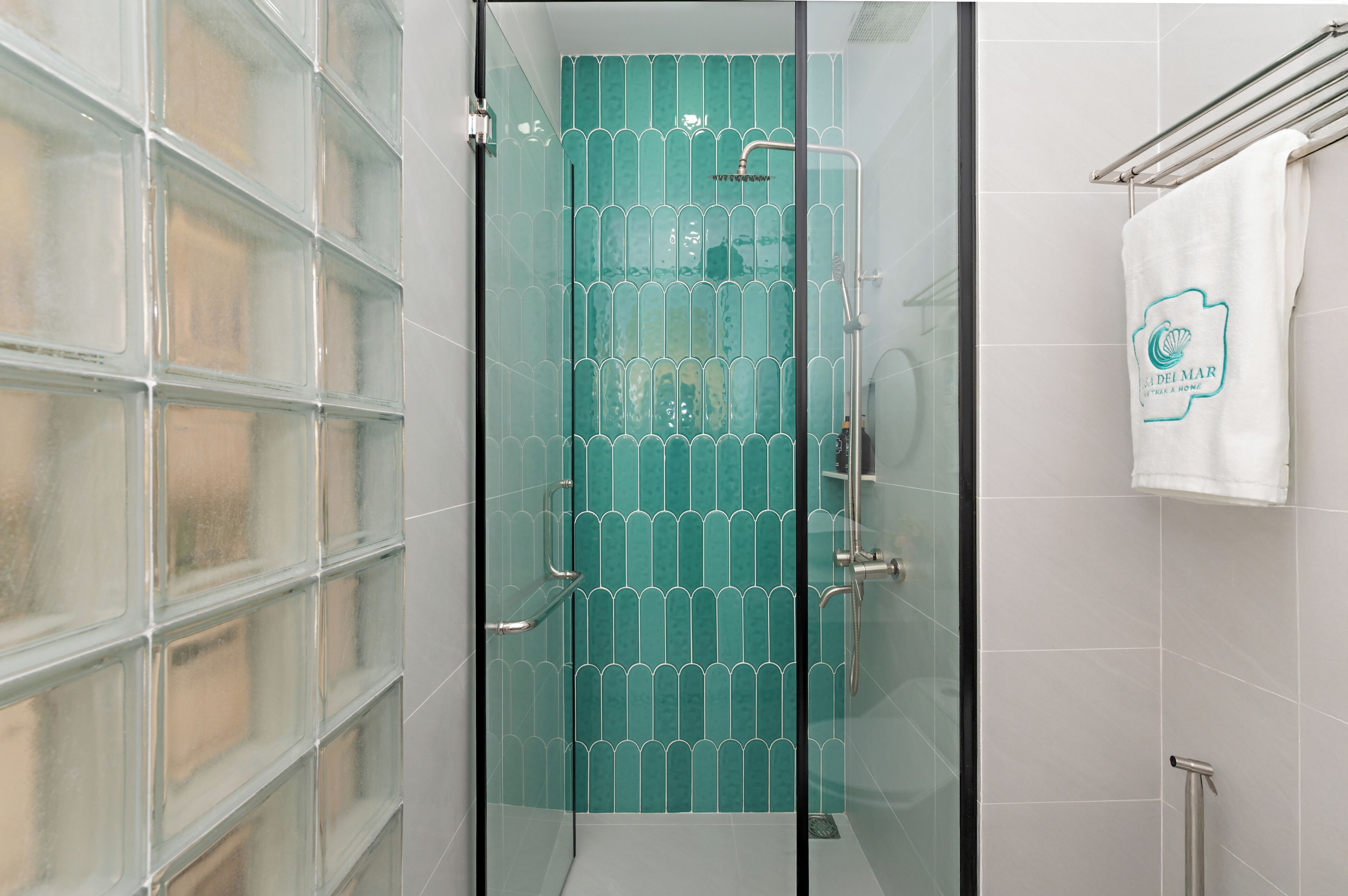 Um chuveiro com paredes de vidro, parede revestida com azulejos verde-água, uma toalha branca em um porta-toalhas e uma porta de vidro.