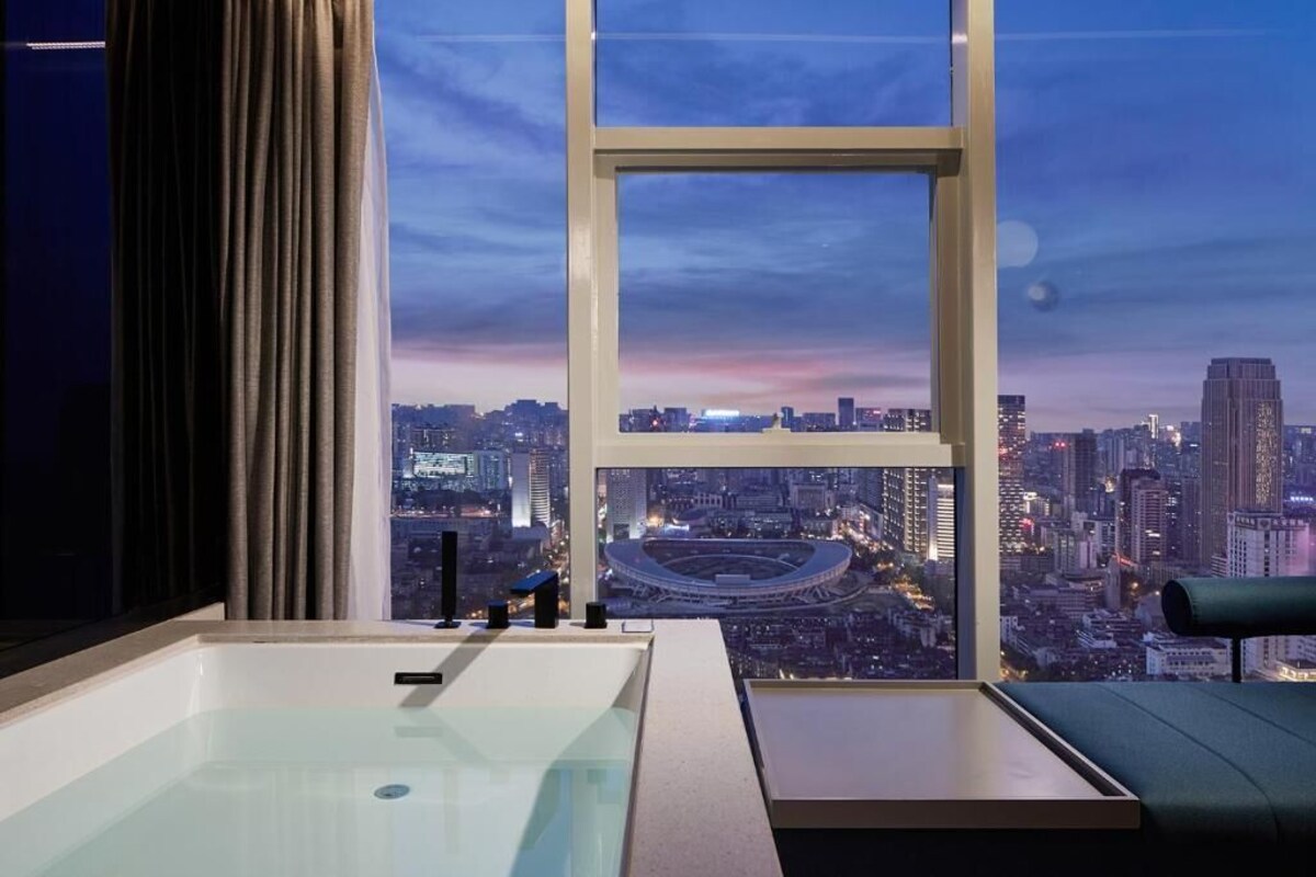 Quarto de hotel moderno com uma janela grande, uma banheira e vista para a paisagem urbana ao entardecer.