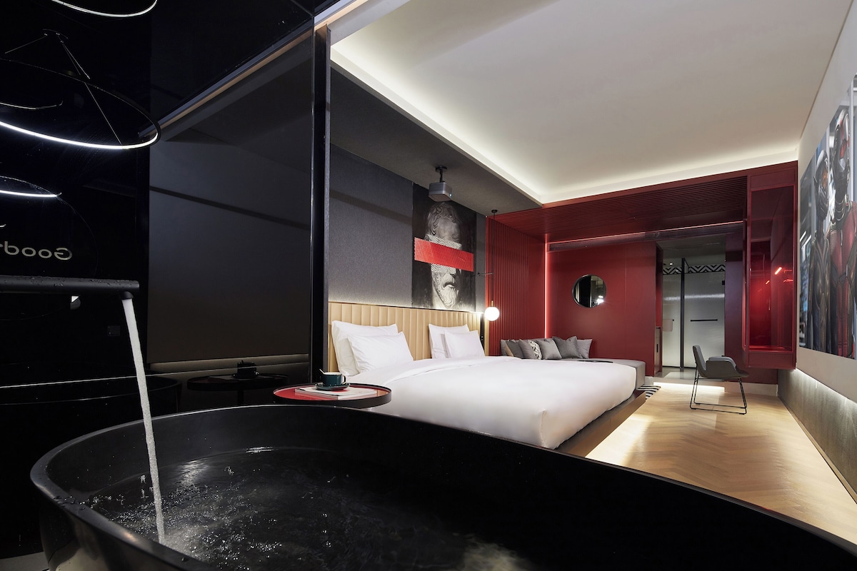 Quarto de hotel moderno com uma banheira grande, cama com roupa de cama branca, parede de destaque vermelha e design minimalista.