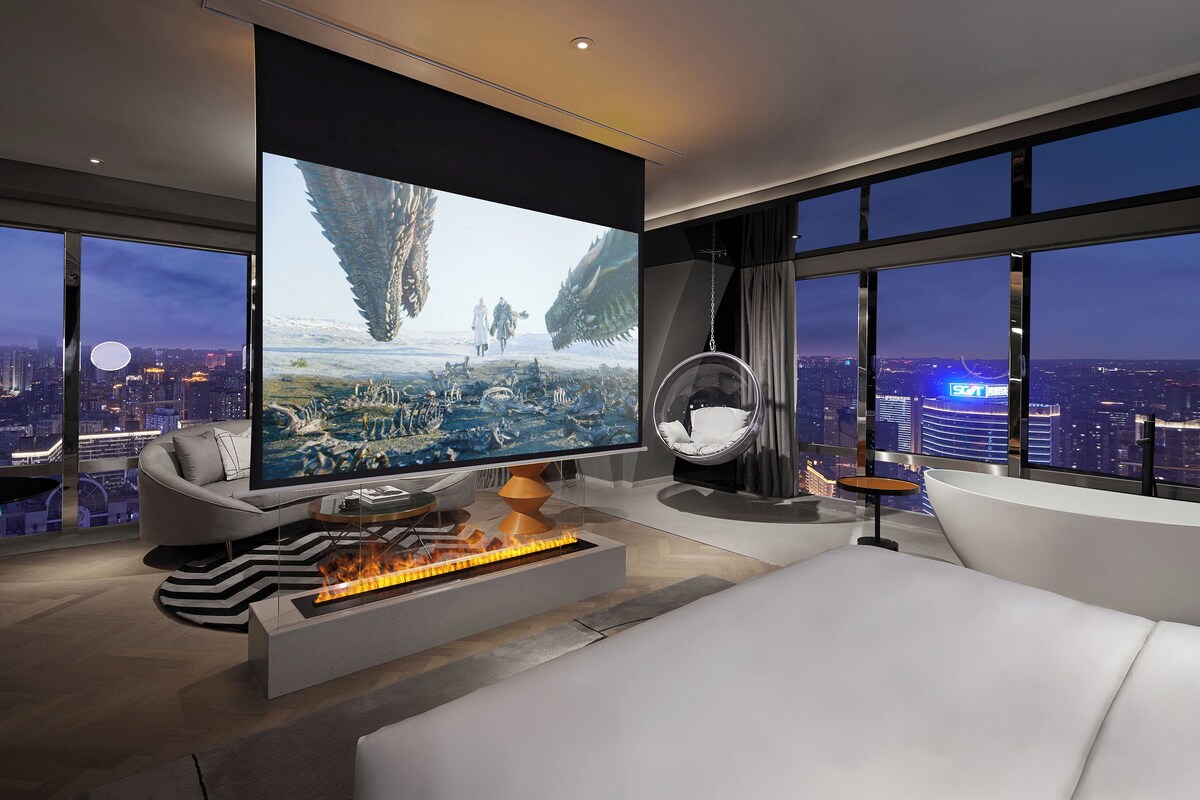 Uma sala de estar moderna com uma televisão de tela plana grande, lareira, sofá e vista para o horizonte da cidade.