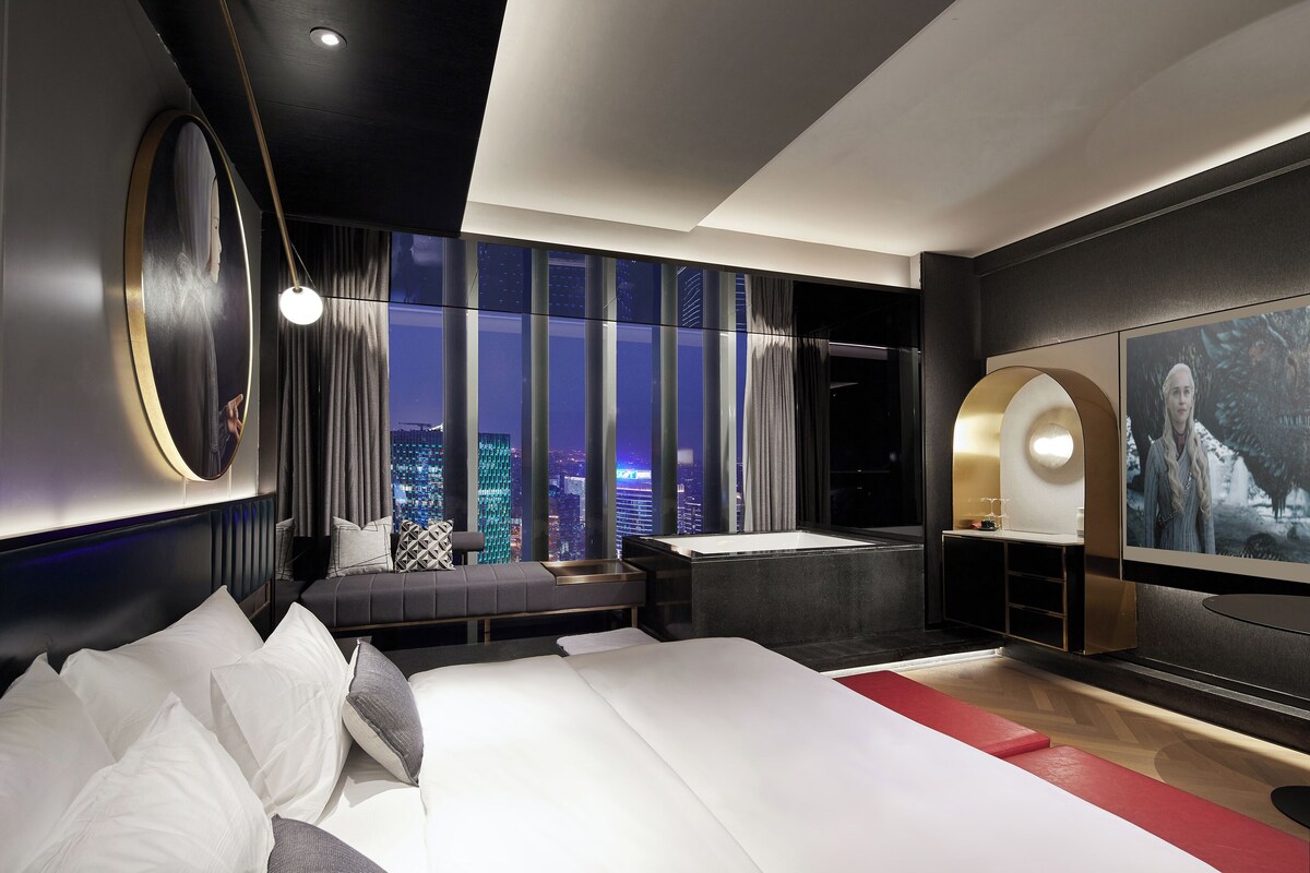 Quarto de hotel moderno com uma cama grande, área de estar, banheira e uma televisão de tela grande.