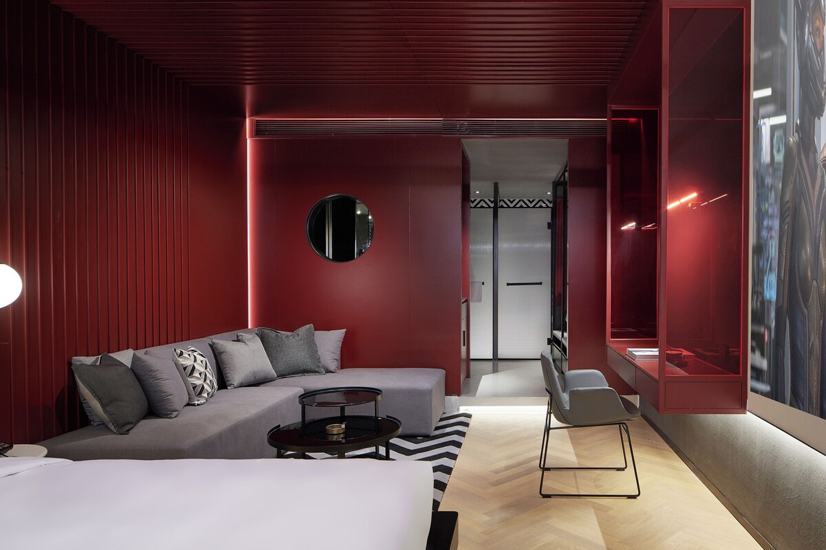 Quarto de hotel moderno com sofá, espelho redondo, mesa pequena, cama e área de estar com uma cadeira.