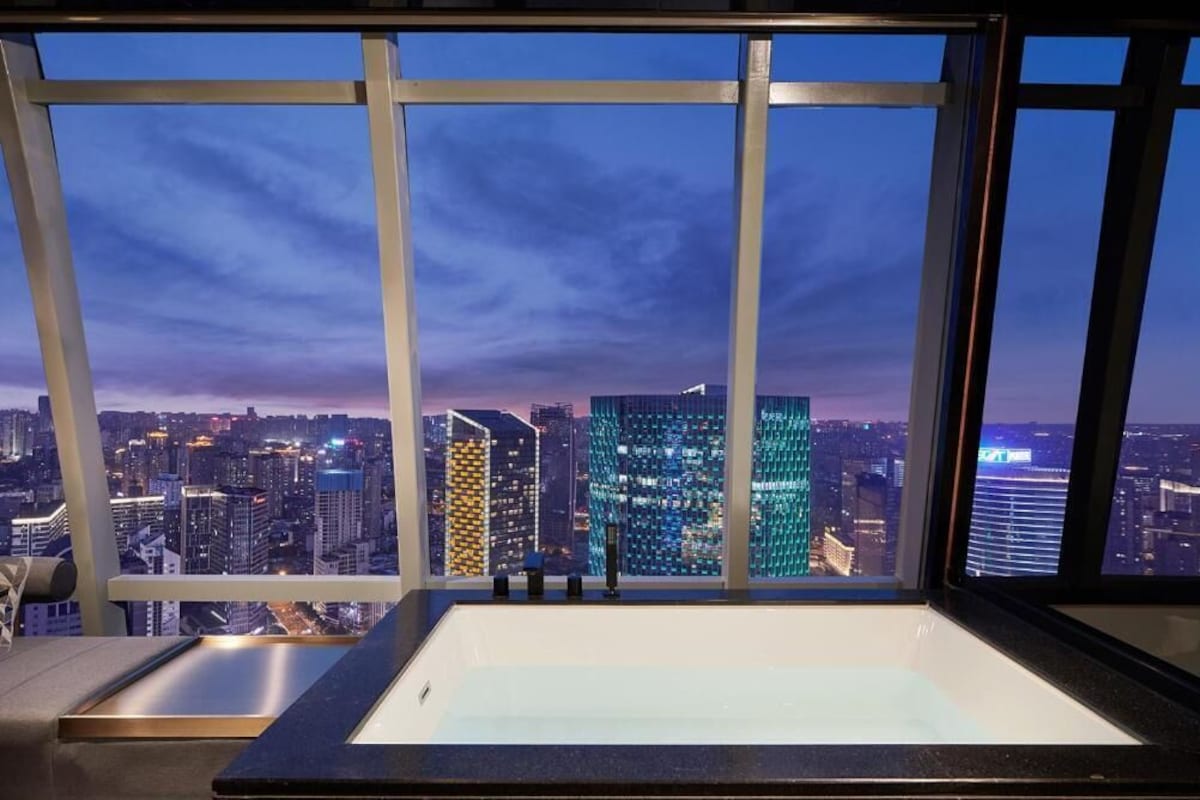 Quarto de hotel moderno com uma grande janela que oferece vista para a cidade ao entardecer.