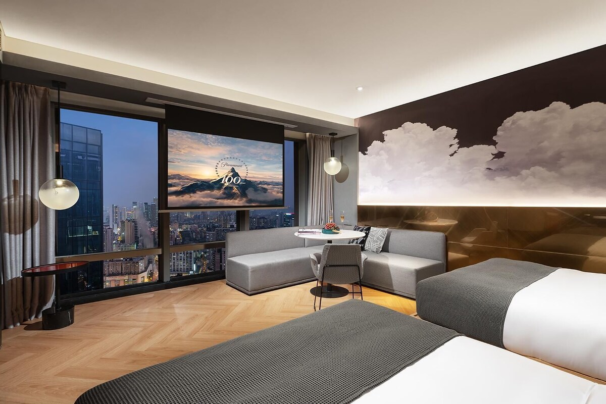Quarto de hotel moderno com vista para a cidade, uma televisão grande, um sofá, uma cama e uma mesa pequena.