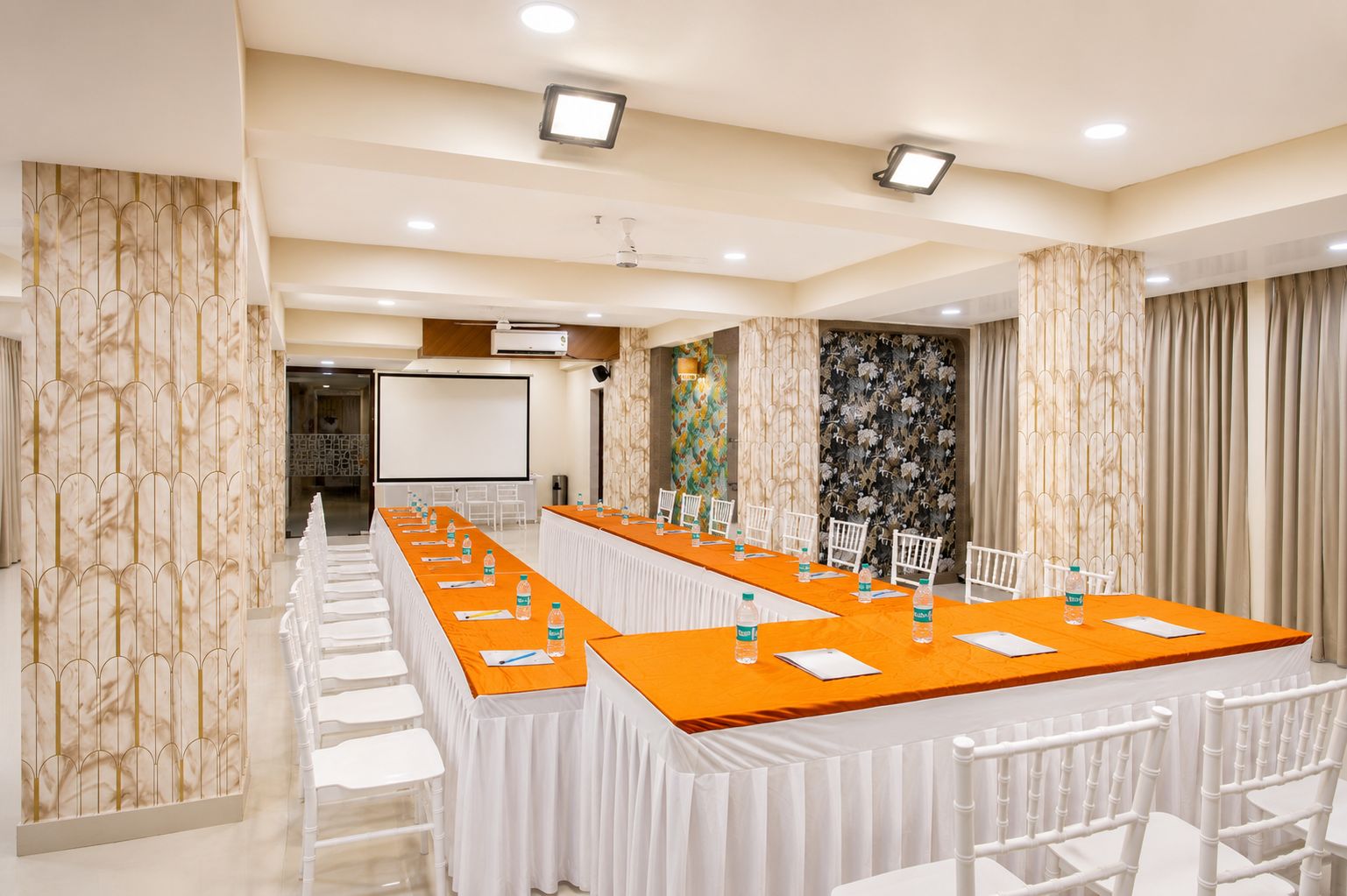 Uma sala de conferências com uma mesa comprida, cadeiras brancas e uma tela de projetor.