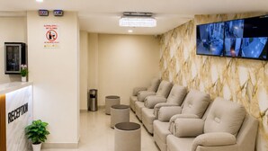 Uma área de recepção com parede de mármore, balcão de recepção, espaço de estar com poltronas de massagem, lixeira e câmera de segurança.