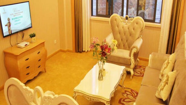 Um quarto com televisão sobre um armário de madeira, uma mesa branca com um vaso de flores, um sofá bege e uma janela grande com cortinas.