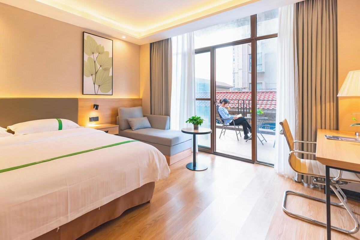 Quarto de hotel com cama, sofá, pequena mesa com planta, cadeira e vista da cidade por grandes janelas.