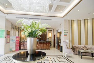 Um saguão de hotel com uma mesa de recepção, um grande vaso decorativo com flores, uma mesa redonda preta e um piso estampado.