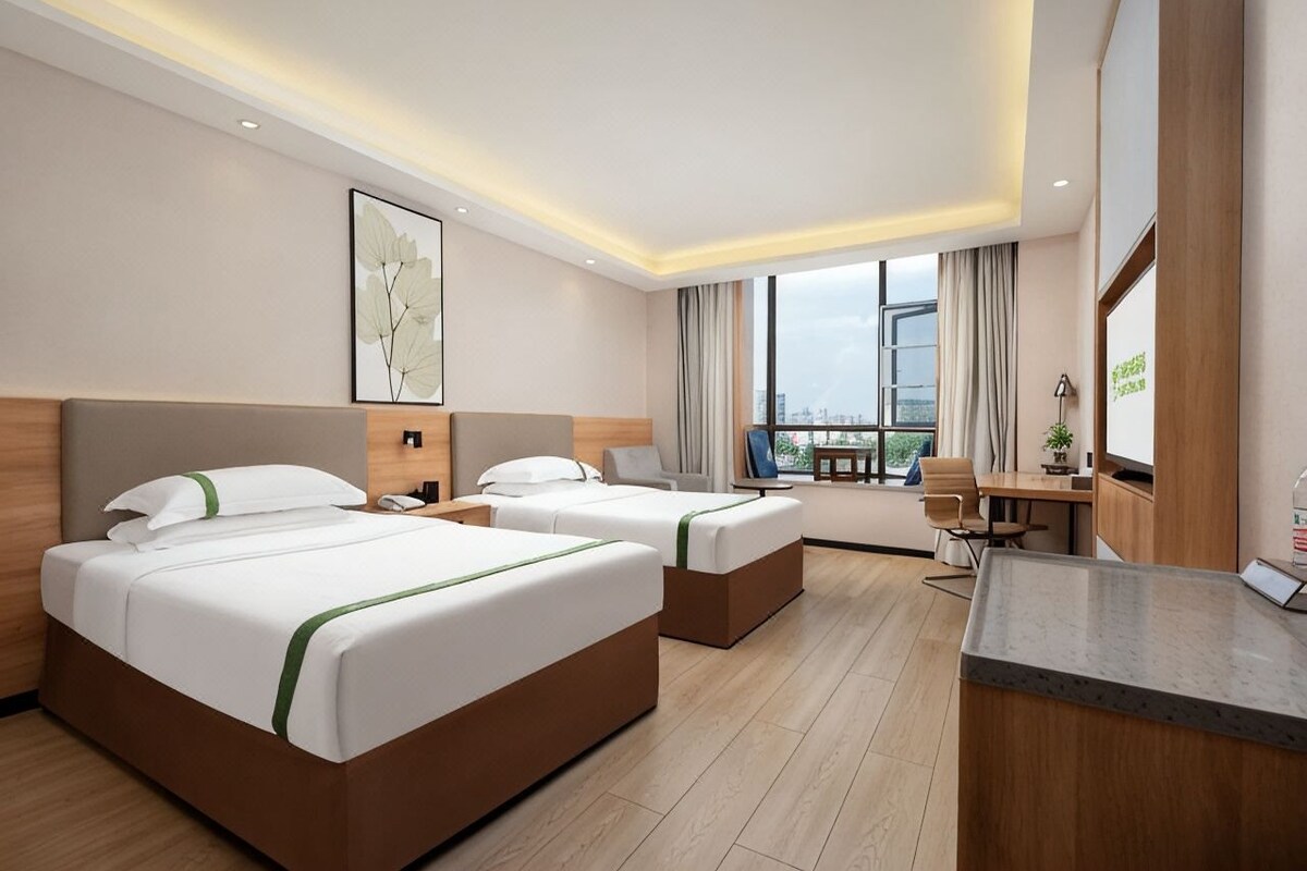 Quarto de hotel com duas camas, uma escrivaninha, uma cadeira e uma janela grande com vista para a cidade.