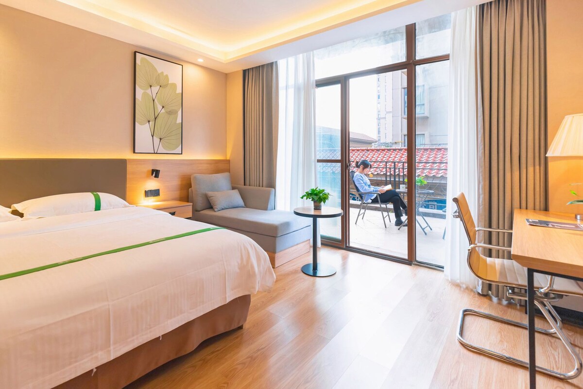 Quarto de hotel com cama, sofá, uma mesinha com planta, cadeira e vista para o exterior.