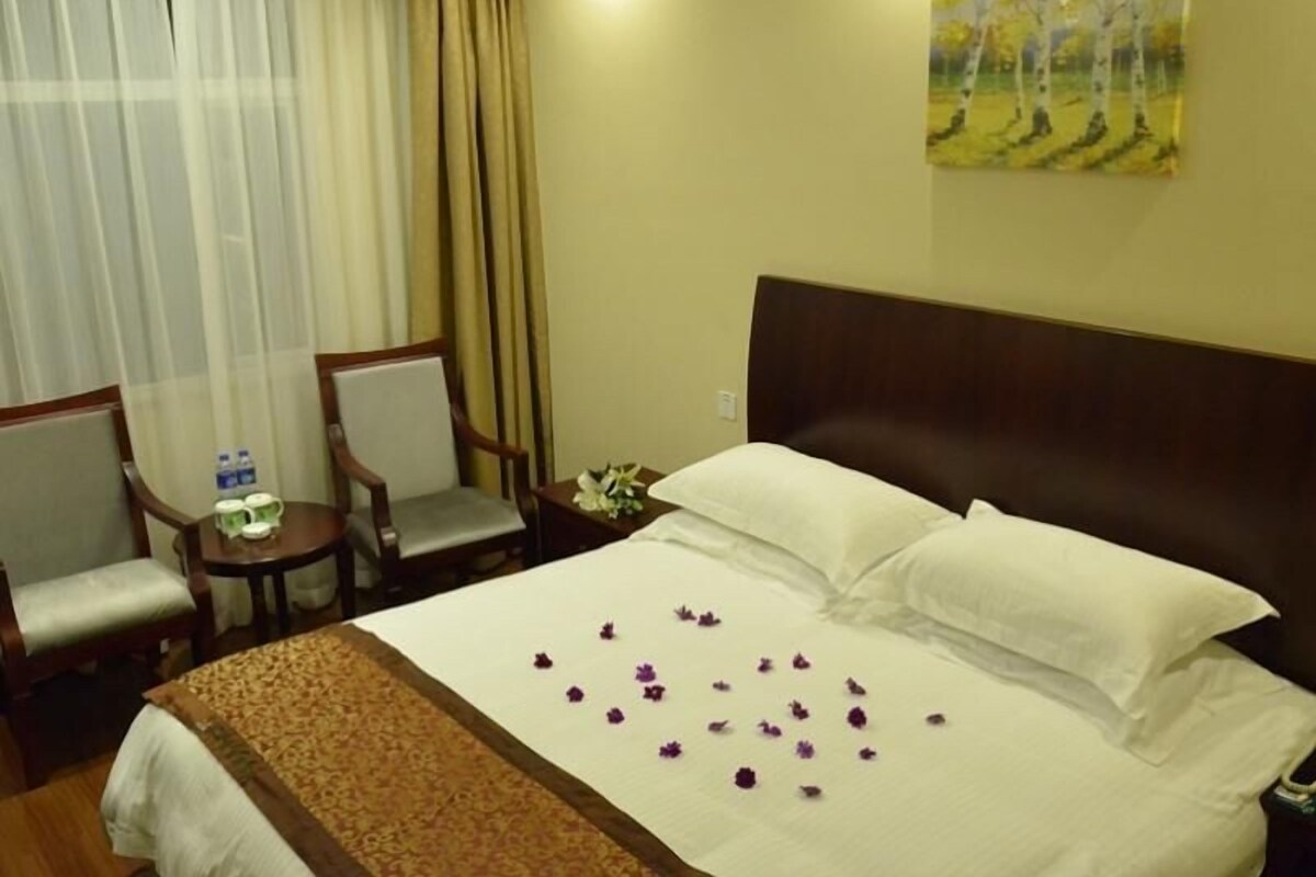 Quarto de hotel com cama, duas cadeiras, uma mesa pequena com uma garrafa de água e um quadro na parede.