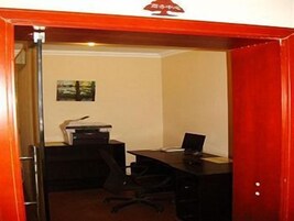 Quarto com escrivaninha, cadeira e computador. Há uma porta com puxador e uma pequena placa acima dela.