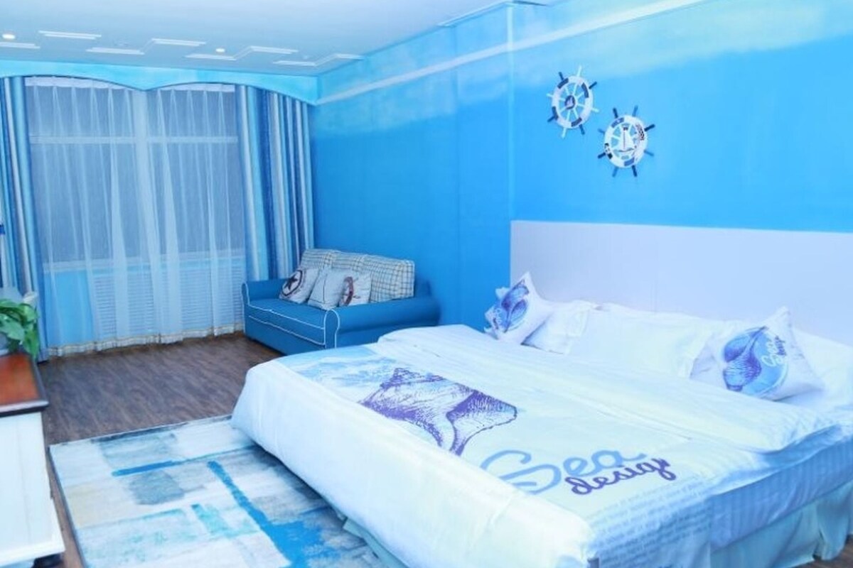 Quarto com cama, sofá azul e janela com cortinas leves.