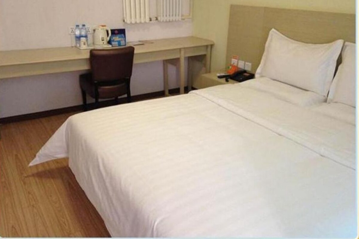 Quarto de hotel com cama, escrivaninha, cadeira, mesa de cabeceira e janela com persianas.