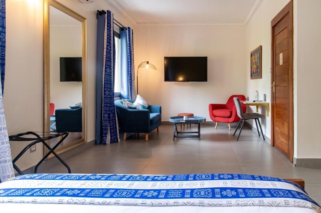 Quarto de hotel com cama, sofá, uma cadeira vermelha, mesa pequena, televisão e porta.