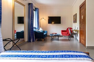 Quarto de hotel com cama, sofá, uma cadeira vermelha, mesa pequena, televisão e porta.