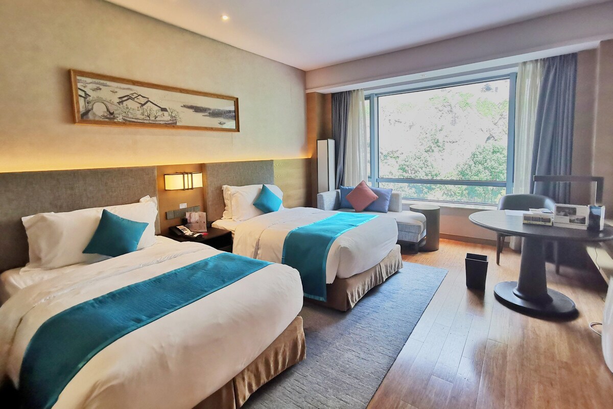 Quarto de hotel com duas camas, uma escrivaninha, uma cadeira, uma mesinha e uma janela grande com vista para árvores.