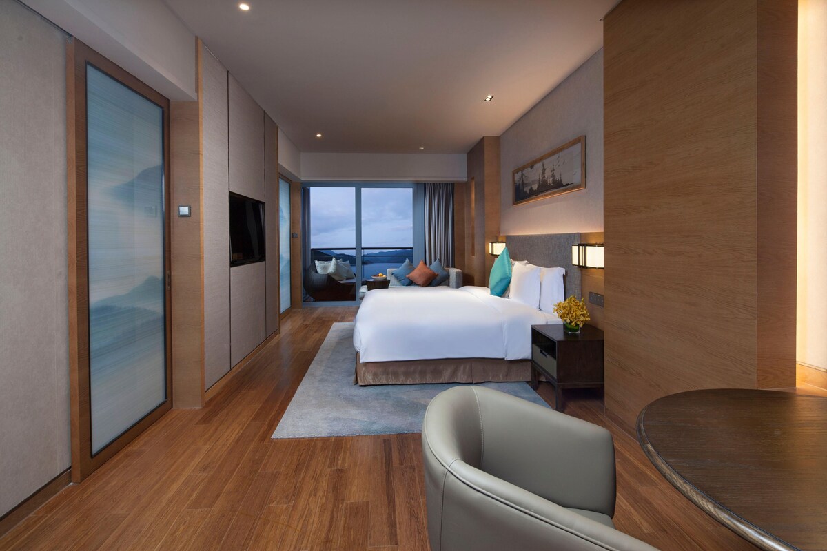 Quarto de hotel moderno com cama grande, área de estar, televisão e vista para o exterior.