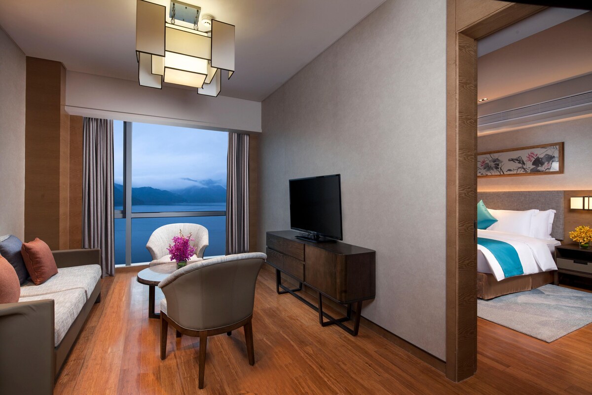 Quarto de hotel com sofá, cadeira, mesa pequena, televisão, cama e uma grande janela com vista.